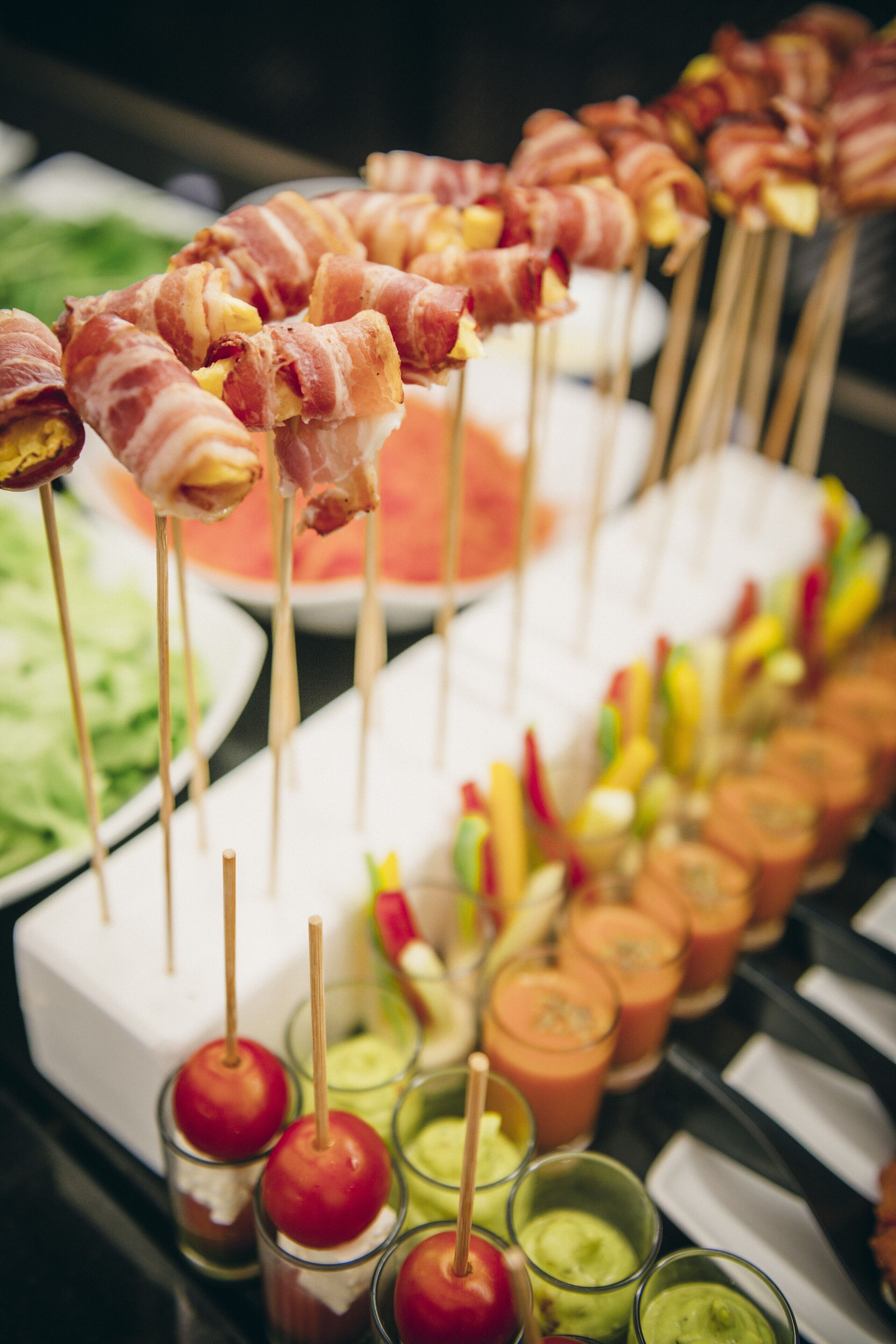Buffet com diversos alimentos, incluindo espetinhos envoltos em bacon, fatias de maçã e diferentes molhos em pequenas taças.