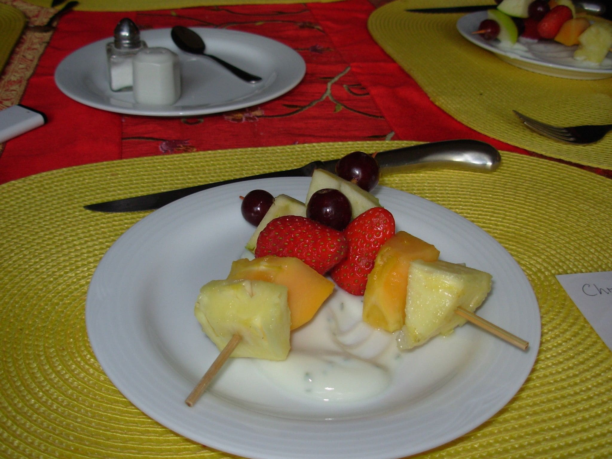 Espetinhos de fruta em um prato branco, com uma colher e um garfo sobre um jogo americano amarelo.