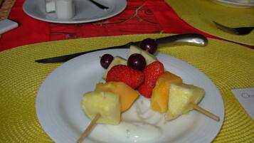 Espetinhos de fruta em um prato branco, com uma colher e um garfo sobre um jogo americano amarelo.