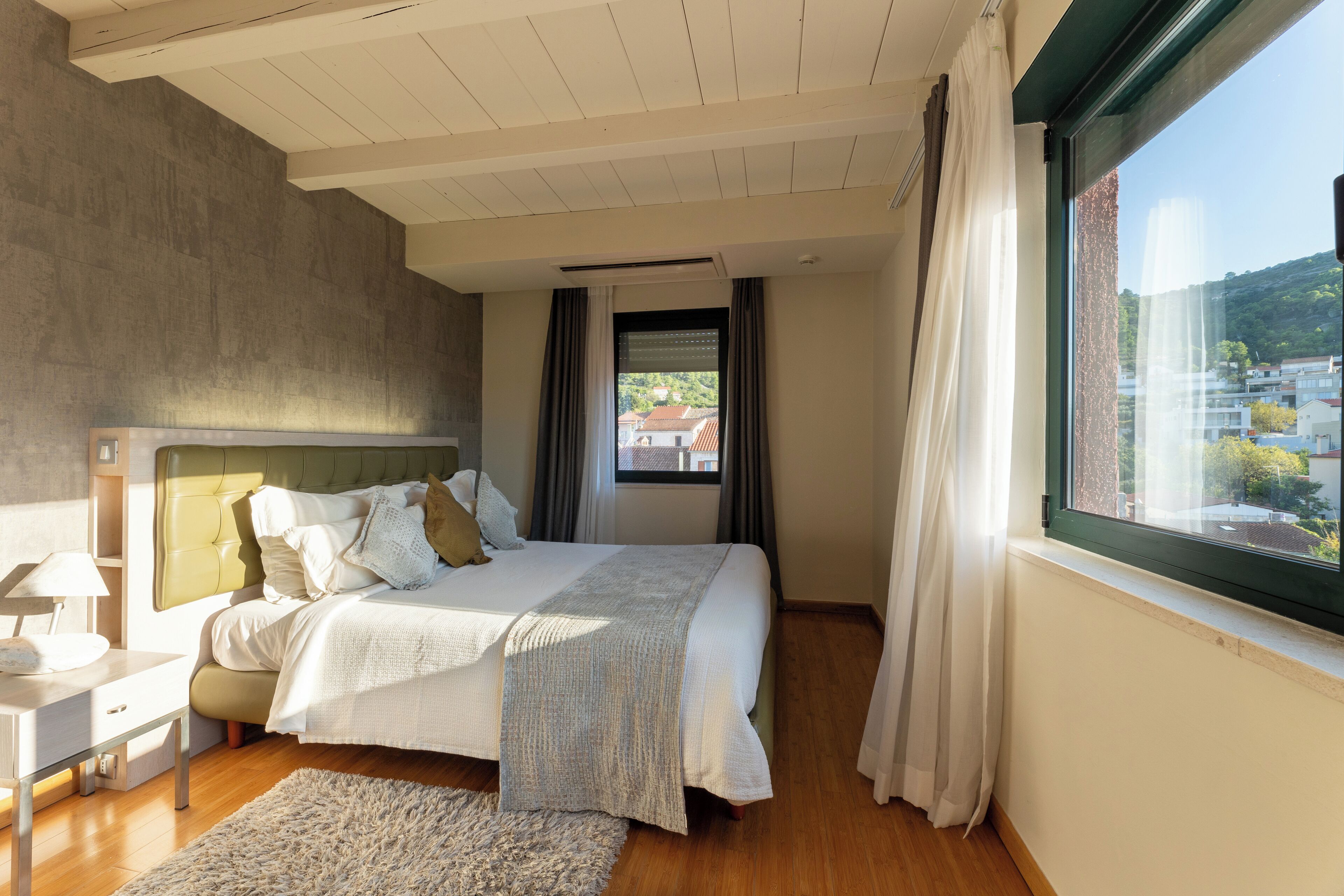 Quarto moderno com uma cama grande, mesa de cabeceira, janela com cortinas e vista para uma área residencial.