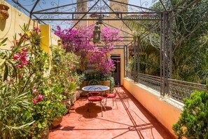 Pátio ao ar livre iluminado pelo sol, com mesa e cadeiras, cercado por plantas exuberantes e um gradeado de ferro.