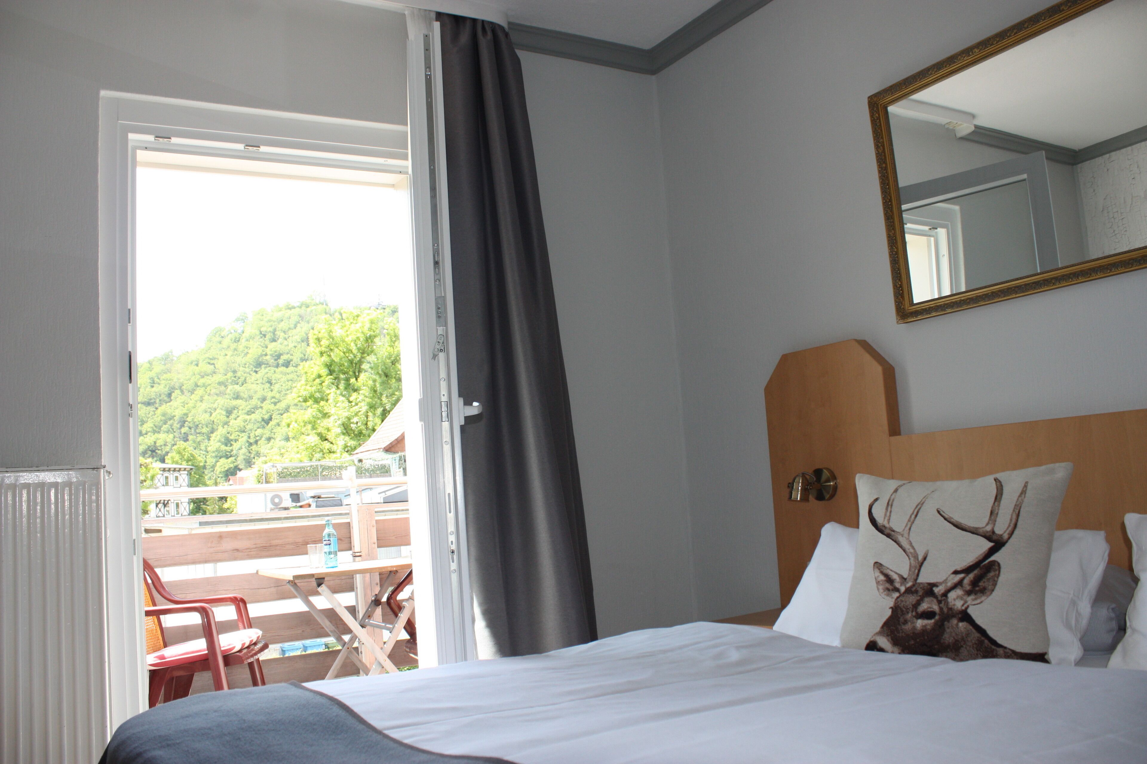 Quarto de hotel com cama, um espelho grande, janela com cortinas e vista para uma varanda com móveis de exterior.