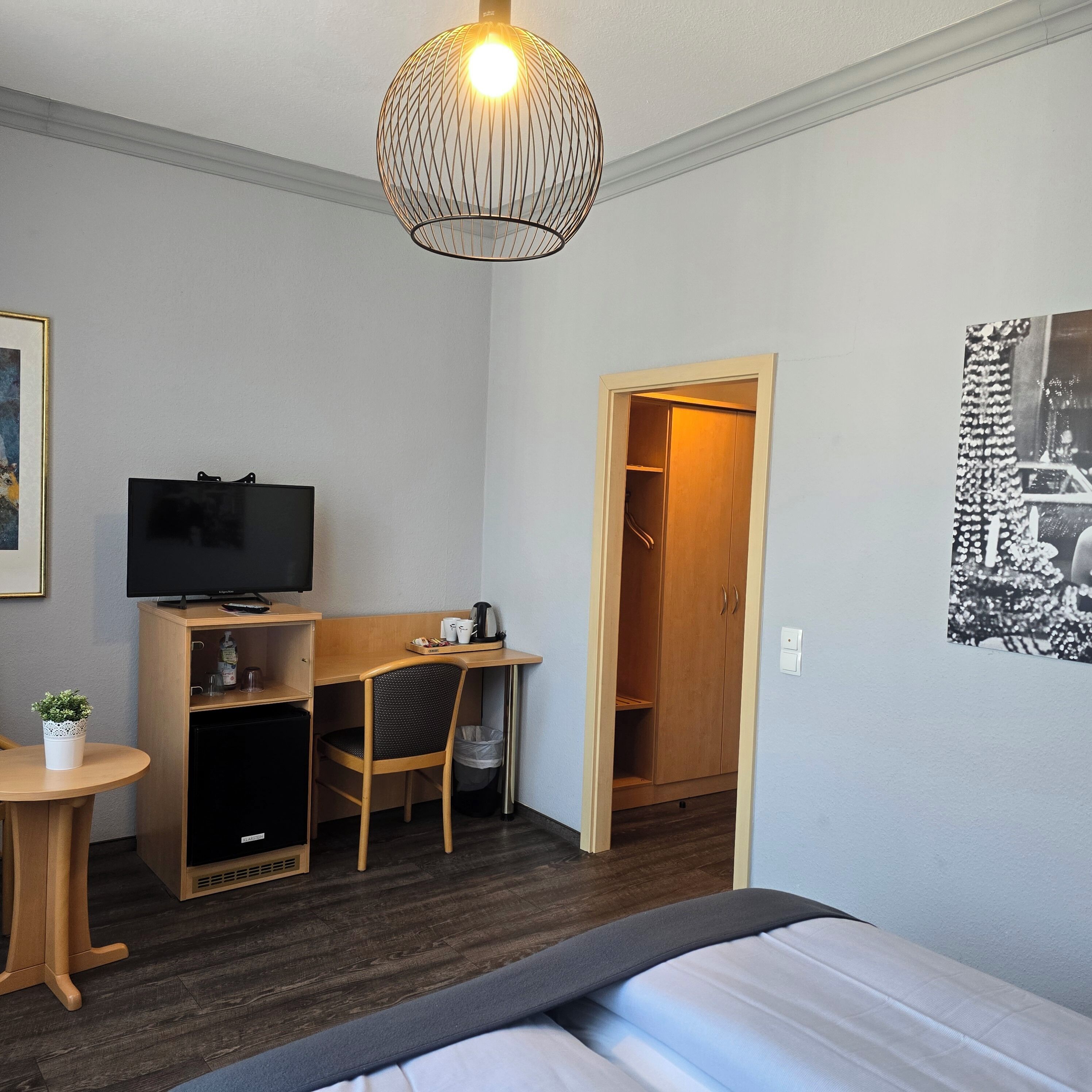 Quarto de hotel com cama, escrivaninha com televisão, mesinha com planta e guarda-roupa.