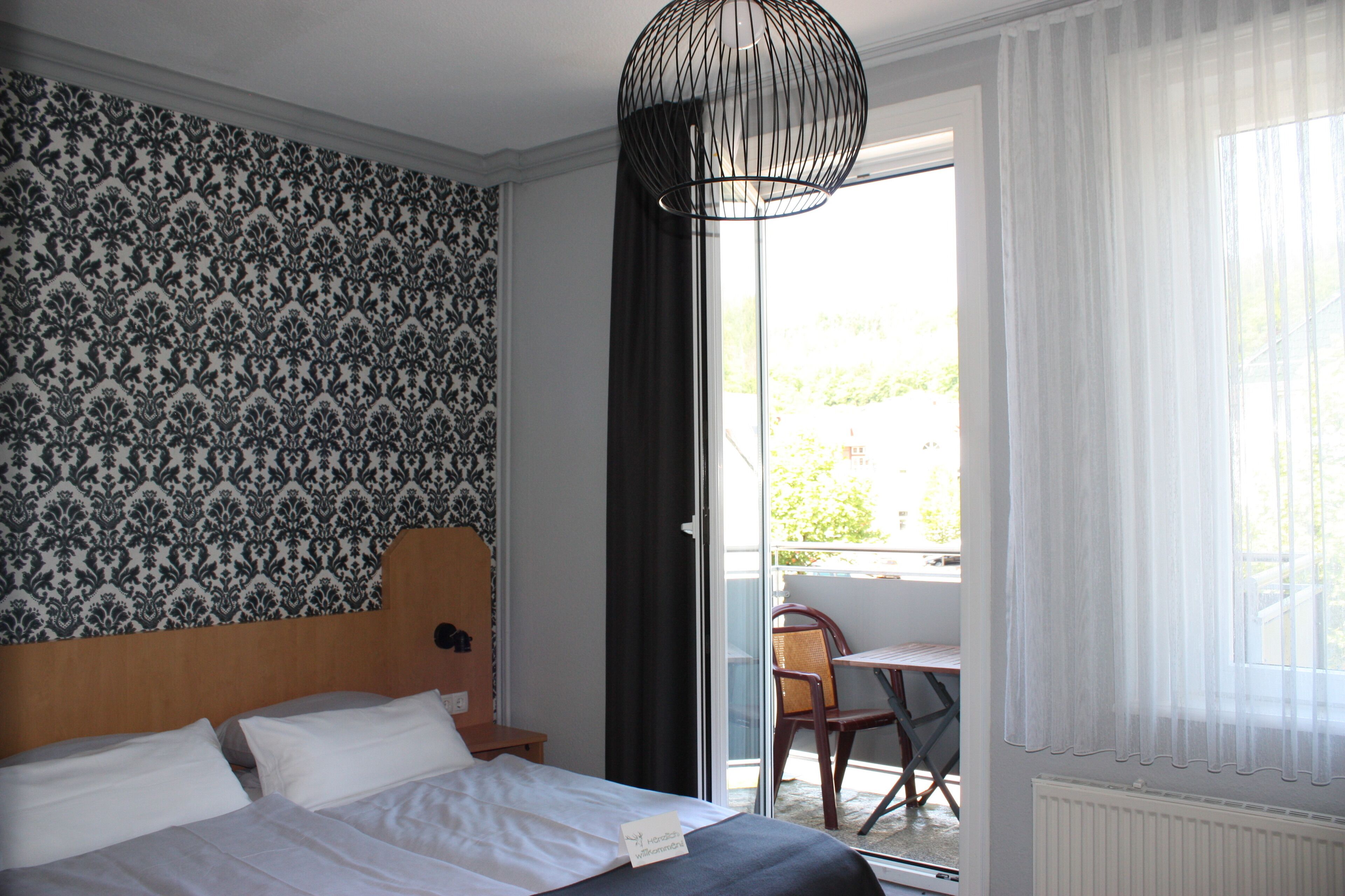 Quarto de hotel com papel de parede estampado, cama com roupa de cama branca, cabeceira de madeira, janela grande com cortinas leves e vista para o exterior.