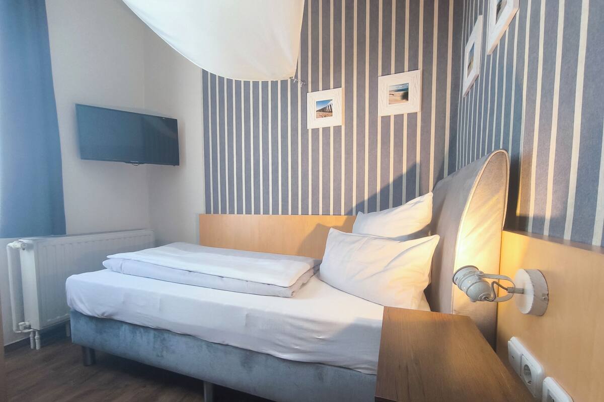 Uma cama de solteiro com cabeceira de madeira, mesa de cabeceira, TV fixada na parede e parede listrada com duas imagens emolduradas.