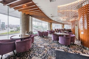 Restaurante moderno com mesas redondas e cadeiras roxas, grandes janelas e um tapete estampado.
