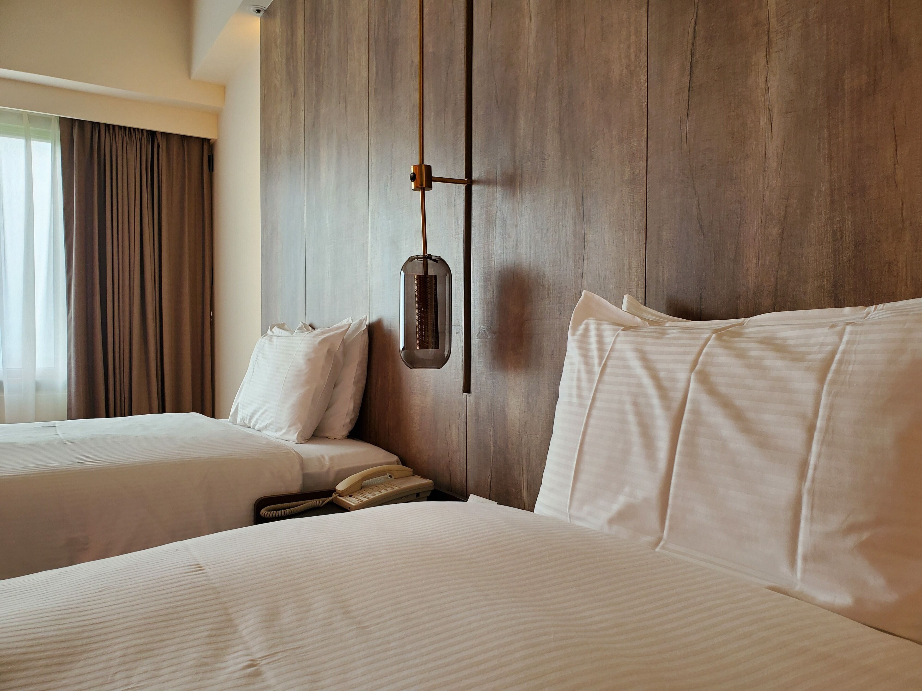 Quarto de hotel com duas camas, cabeceira de madeira, luminária pendente e um telefone.