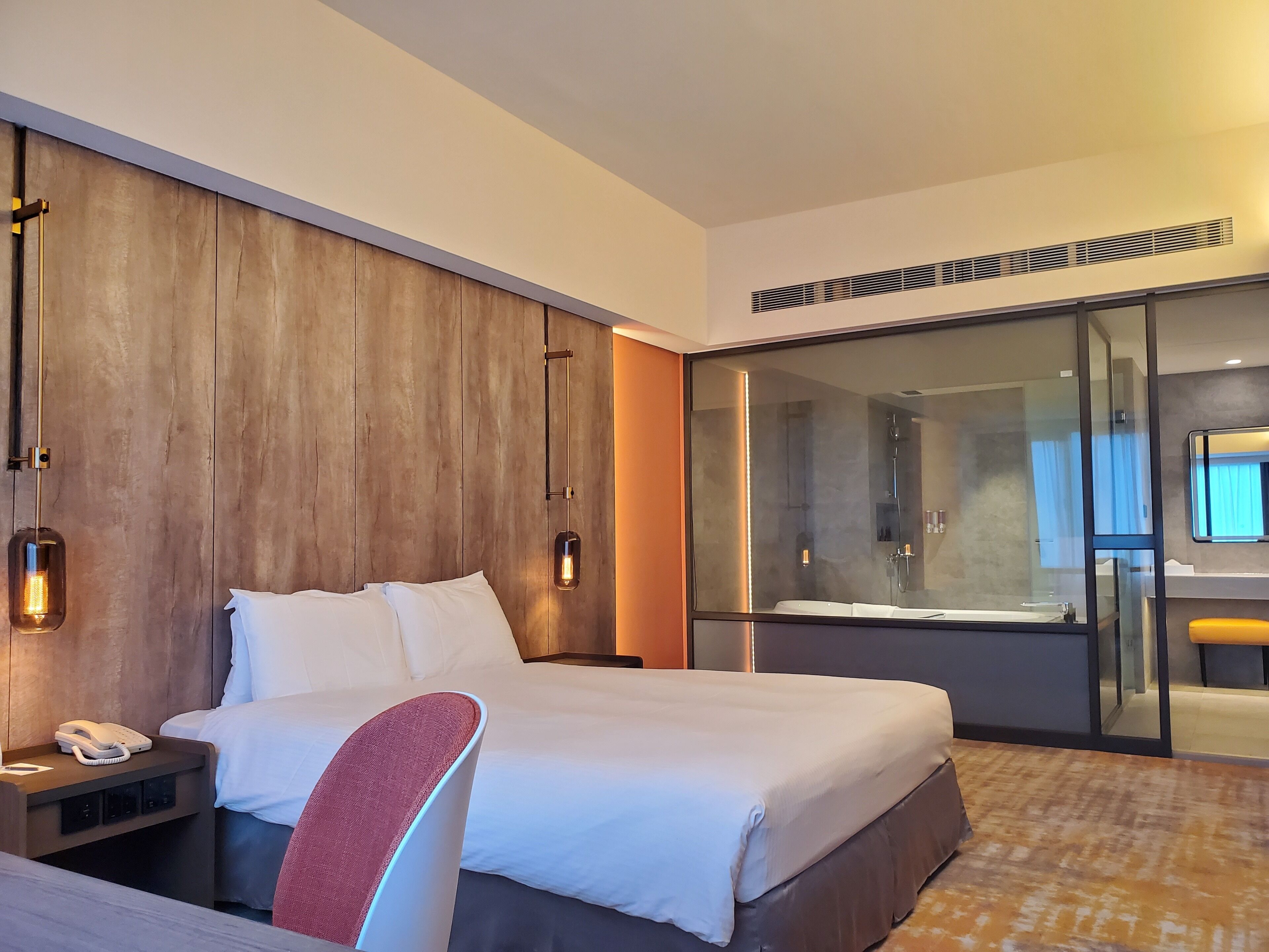 Quarto de hotel moderno com uma cama grande, telefone ao lado da cama, uma cadeira vermelha e um banheiro com banheira, separado por vidro.