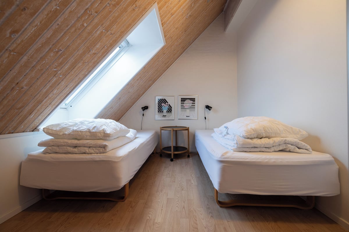 Um quarto de sótão pequeno e acolhedor, com duas camas simples, um claraboia e uma pequena mesa com duas luminárias fixadas na parede.