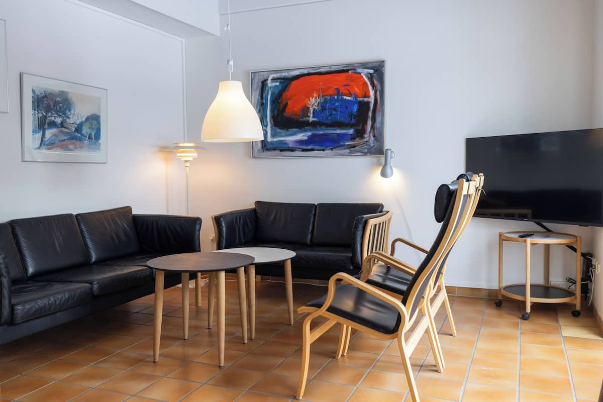 Uma sala de estar moderna com um sofá de couro preto, uma mesa de centro de madeira, uma poltrona de couro preto, uma mesinha lateral redonda, um abajur, um quadro e uma TV de tela plana.