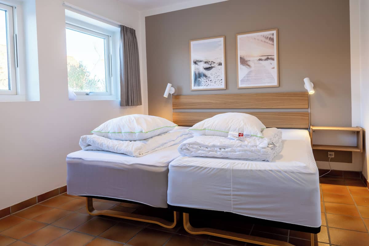 Uma cama de casal com roupa de cama branca, cabeceira de madeira, duas luminárias fixadas na parede e dois quadros enquadrados.