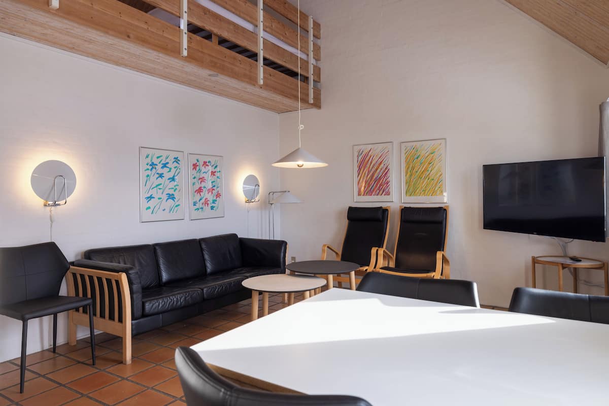 Uma sala de estar moderna com um sofá de couro preto, cadeiras de madeira, uma mesa branca e uma televisão de tela plana.