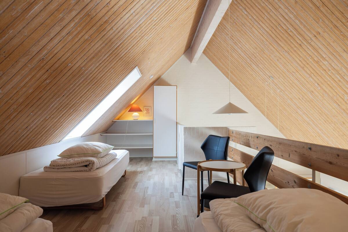 Um quarto de sótão acolhedor, com teto inclinado, uma cama, uma cadeira e uma pequena mesa.