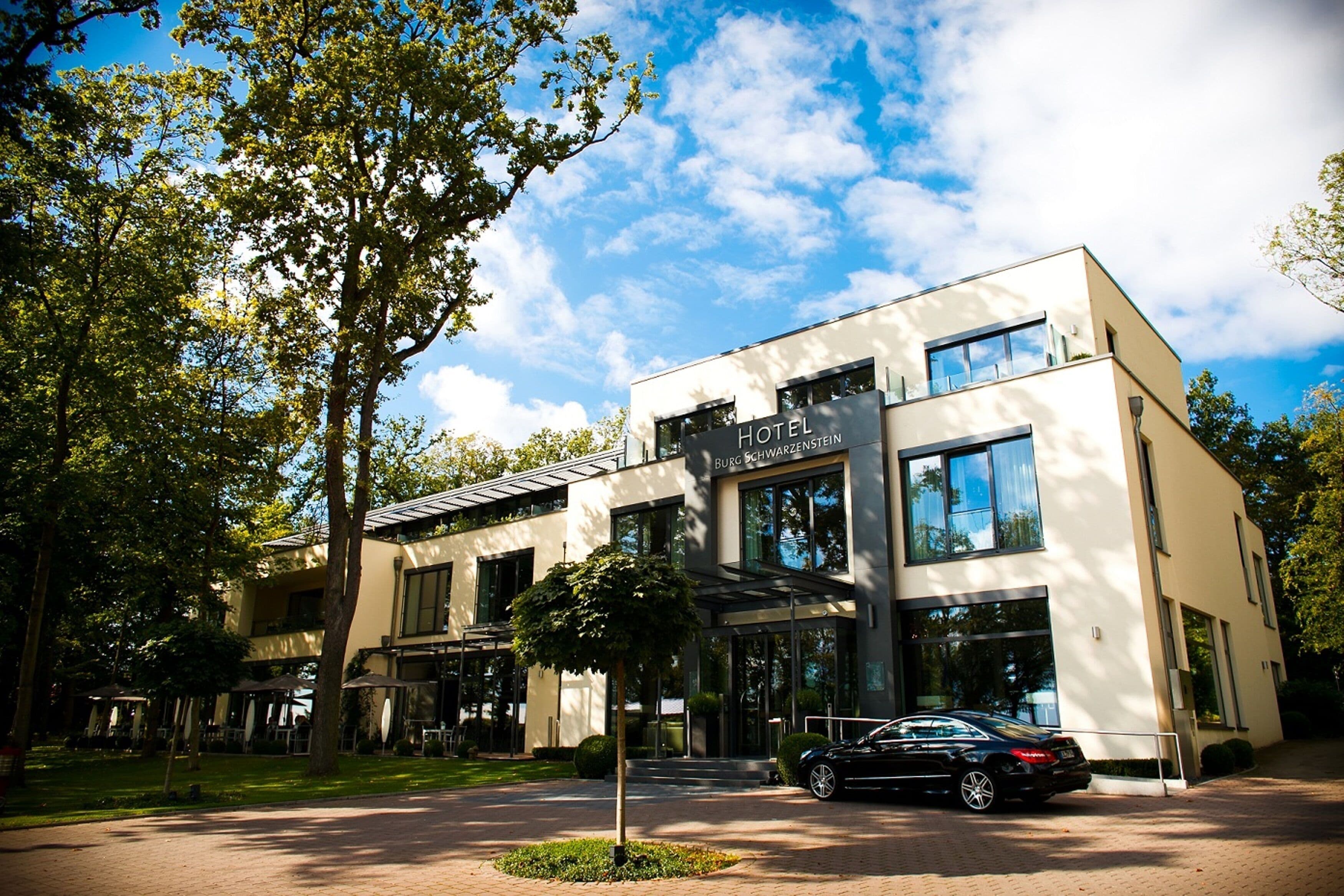 Hotel moderno com entrada visível, um carro estacionado em frente e área externa bem cuidada com árvores.
