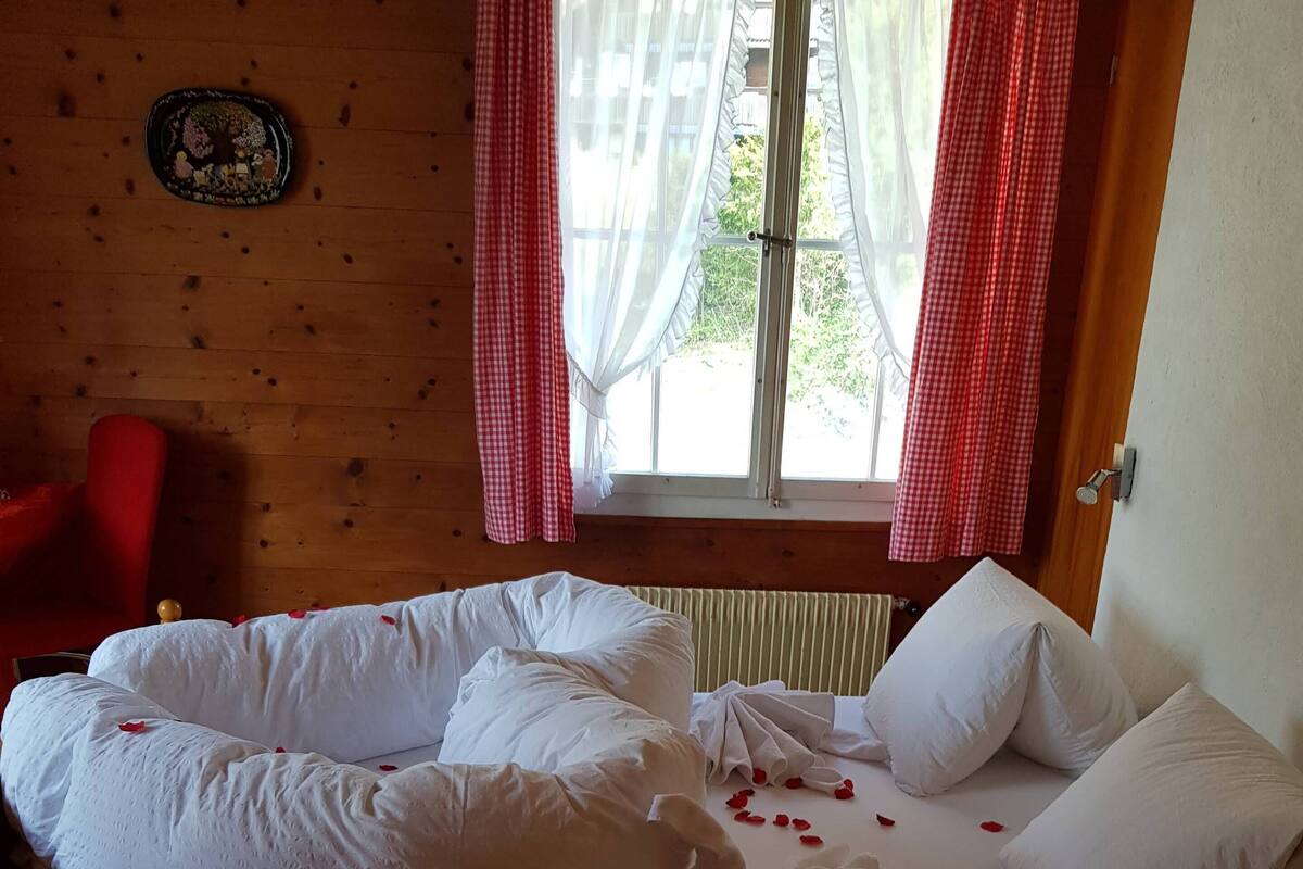 Um quarto aconchegante com piso de madeira, uma cama com roupa de cama branca e p étalas de rosa, uma cadeira vermelha e uma janela com cortinas vermelhas.