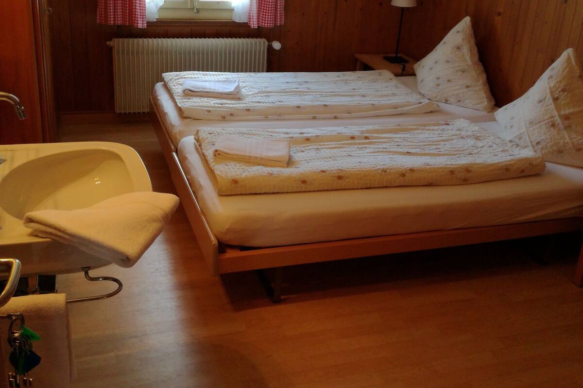 Quarto com duas camas, piso de madeira, janela com cortinas vermelhas, radiador e pia.
