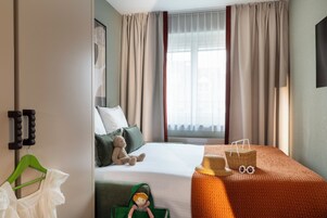 Quarto de hotel com cama, cabide verde, um ursinho de pelúcia, uma bolsa verde e uma cesta de vime com óculos de sol.