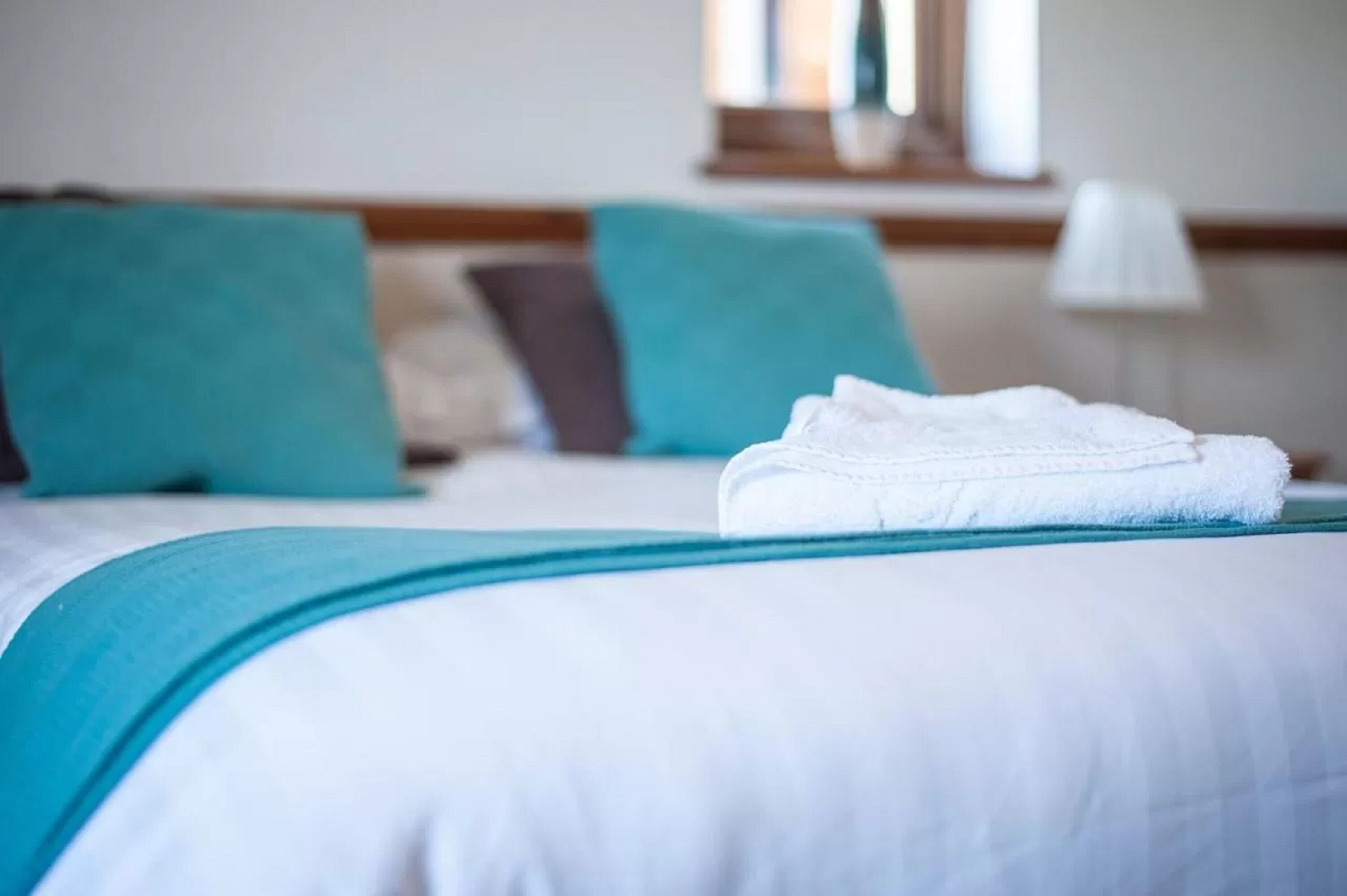 Cama bem arrumada com colcha branca e azul-turquesa, toalha branca dobrada e almofadas azul-turquesa.