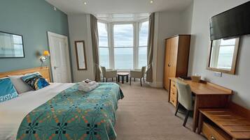 Quarto de hotel com cama, escrivaninha, cadeira, televisão e vista para o mar.