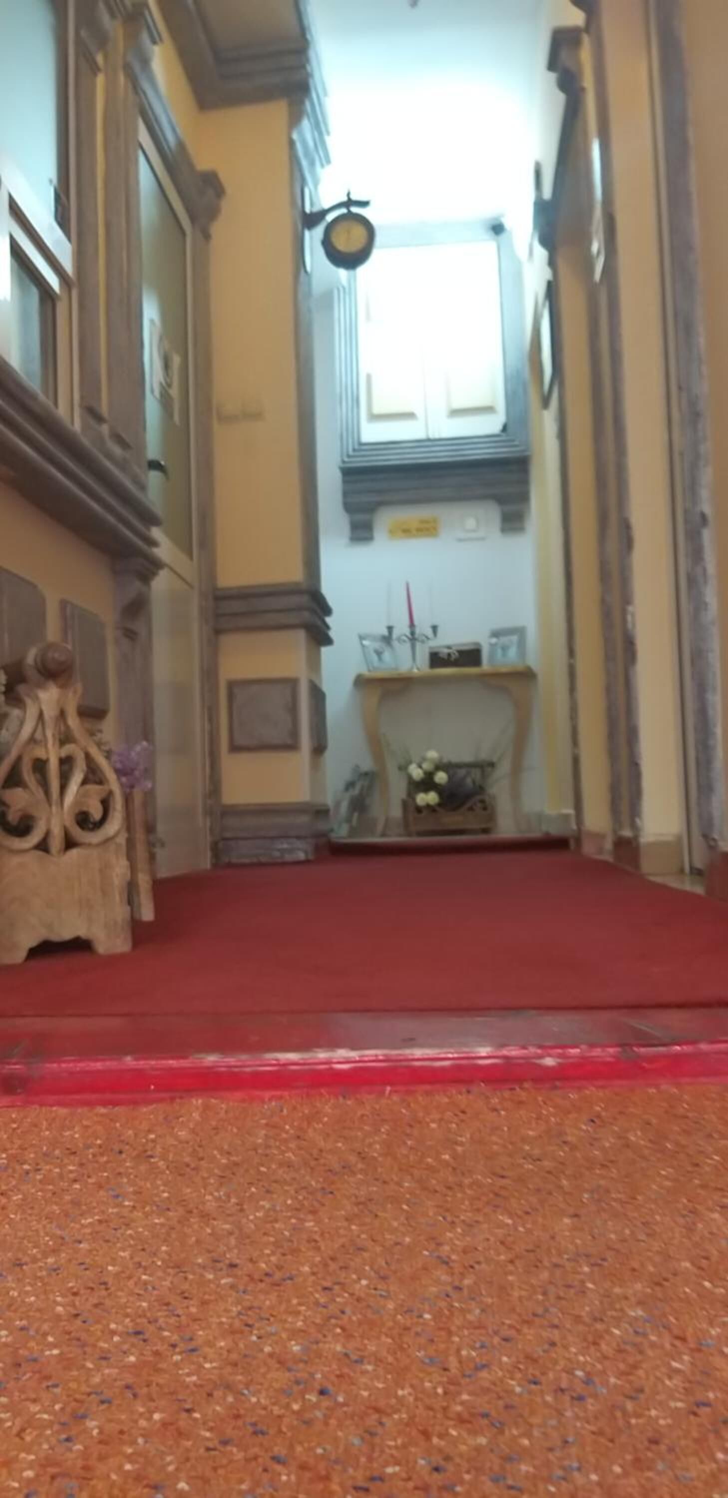 Um corredor estreito com carpete vermelho que leva a um pequeno altar com vela e flores.