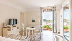 Quarto de hotel com cama, televisão, escrivaninha com computador, cadeira, mesinha e varanda com plantas.