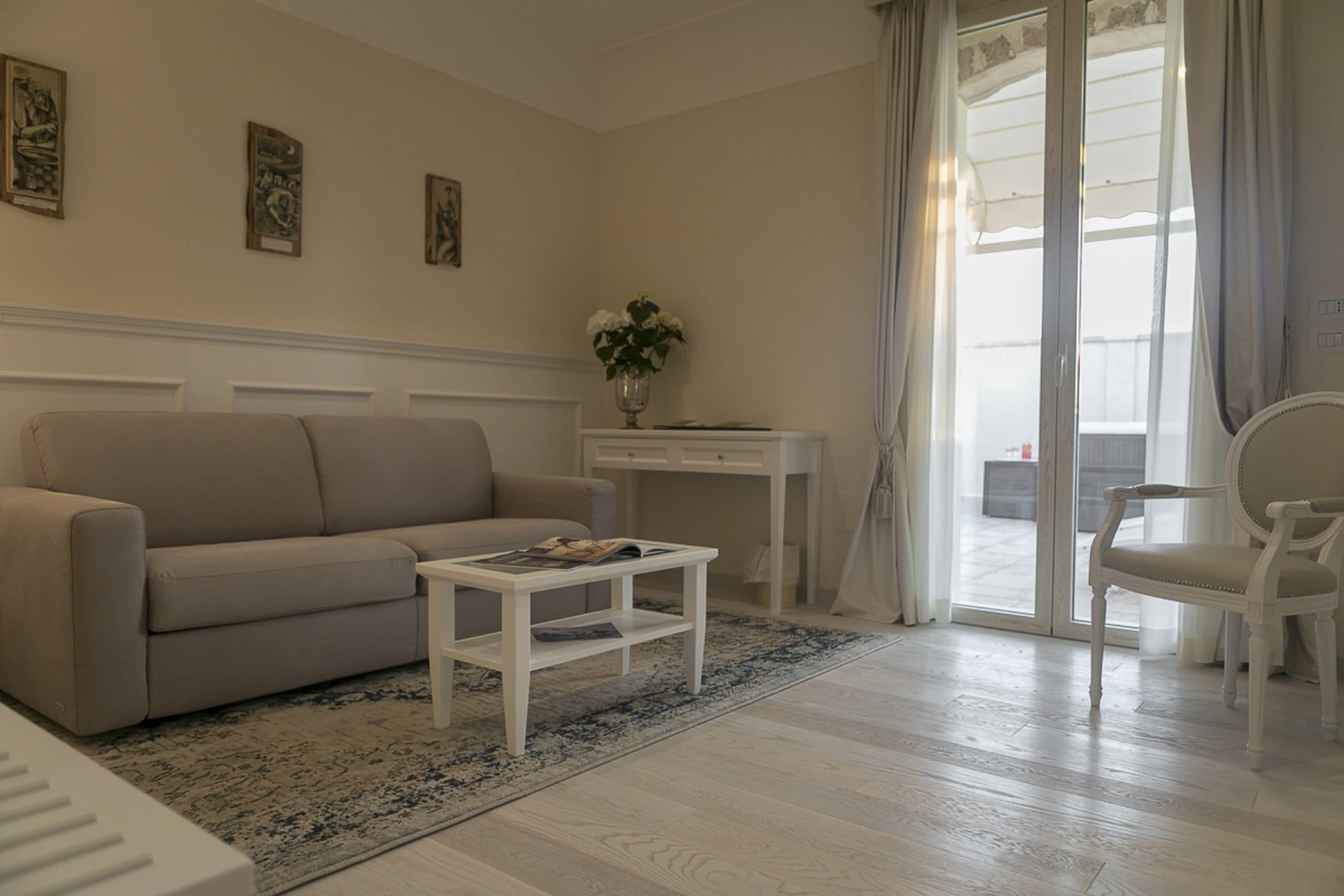 Uma sala de estar com um sofá bege, uma mesa de centro branca e uma cadeira branca. Há uma pequena mesa branca com um vaso de flores. O quarto tem uma janela grande com cortinas.