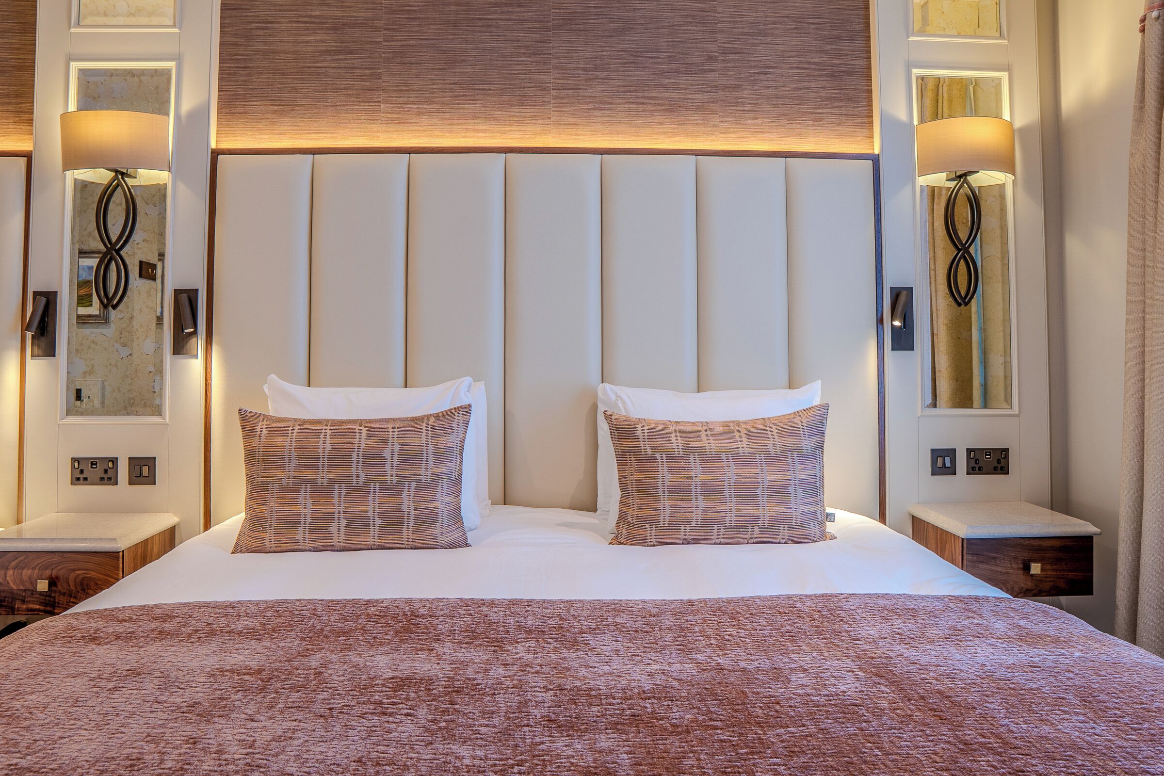 Quarto de hotel com uma cama grande, duas mesas de cabeceira, uma cabeceira com painéis verticais e luminárias fixadas na parede.