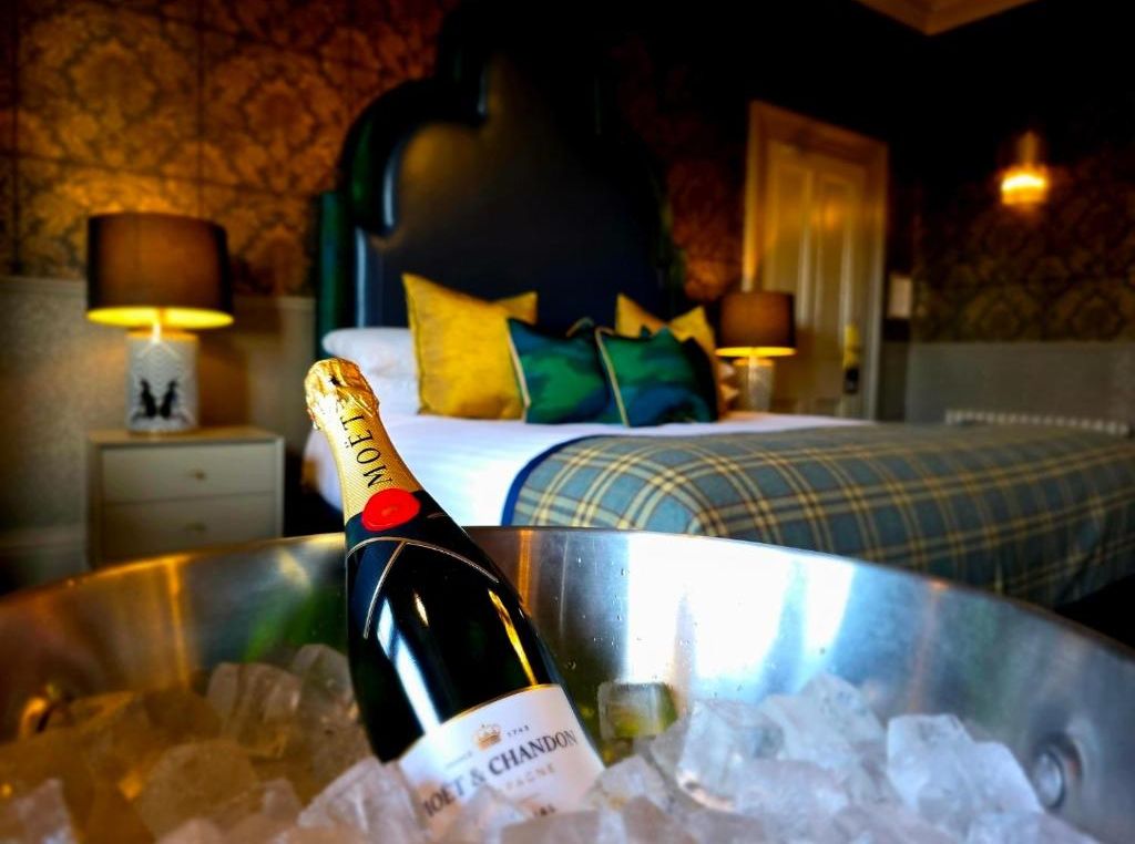 Uma garrafa de champanhe Moët & Chandon num balde de metal cheio de cubos de gelo, colocada num quarto com cama, almofadas e candeeiros.