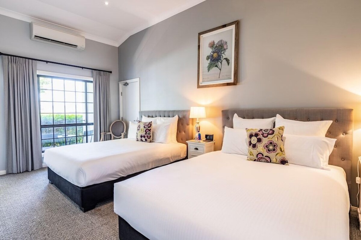 Quarto de hotel com duas camas, uma janela grande com cortinas, um quadro na parede e uma mesa de cabeceira com abajur.