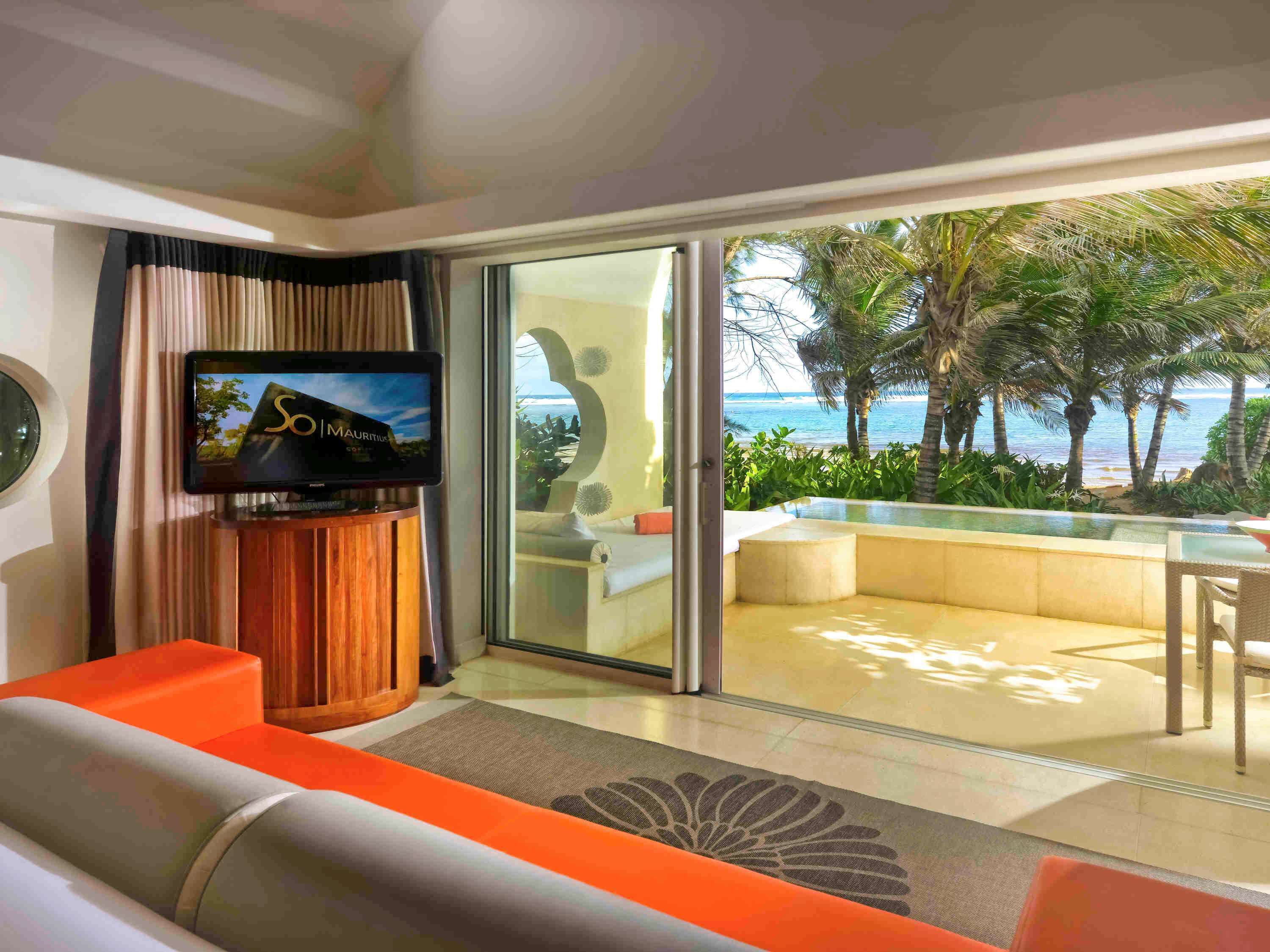 Sala de estar moderna com televisão exibindo 'So Mauritius' e vista para o oceano através de grandes portas de vidro.