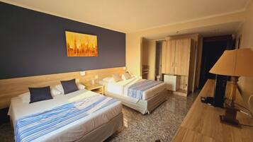 Quarto de hotel com duas camas, um quadro na parede, uma escrivaninha com abajur e um guarda-roupa.