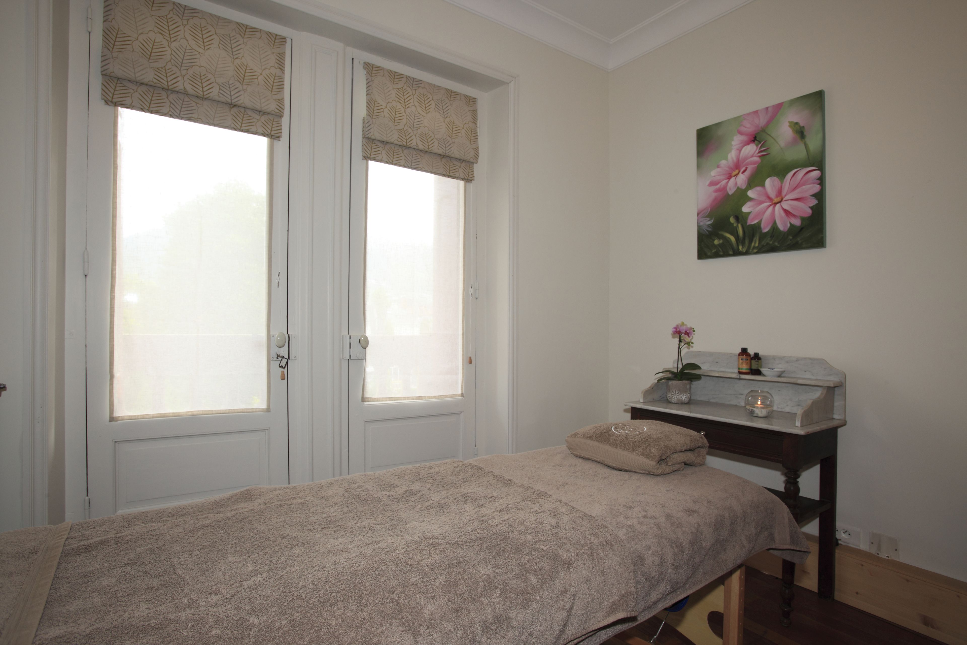 Quarto com cama, mesa de cabeceira com um vaso e uma garrafa, janela com persiana estampada de folhas e um quadro de flores na parede.