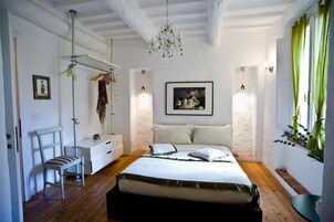 Um quarto com cama, cadeira, mesa de cabeceira, planta pendente e um quadro na parede.