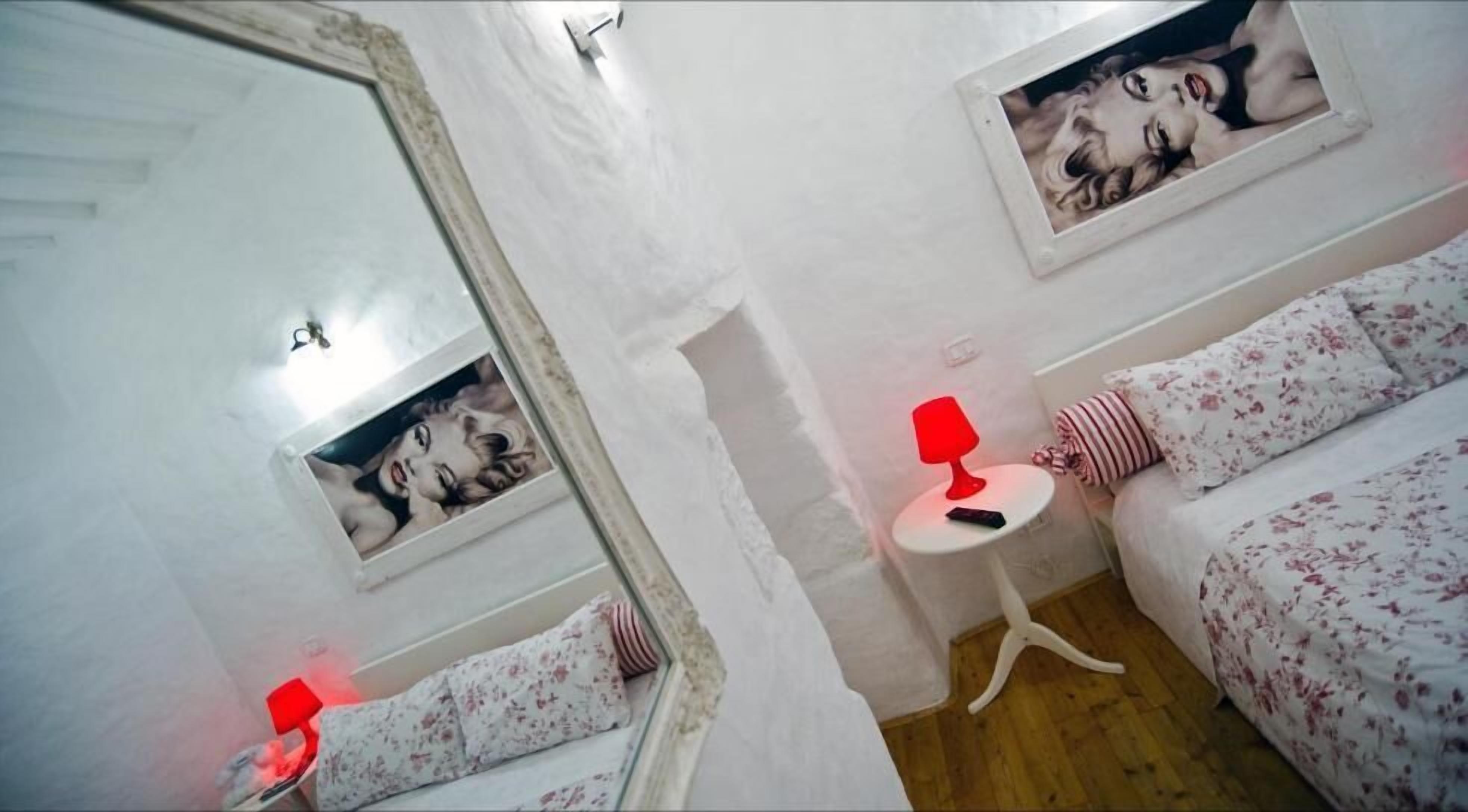 Quarto com um espelho grande, mesa de cabeceira com abajur vermelho e colcha estampada com flores.