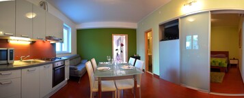 Cozinha compacta com eletrodomésticos modernos, área de jantar com mesa de vidro e um quarto com cama e mesa de cabeceira.