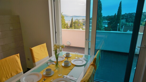 Uma sala de jantar com mesa posta para quatro pessoas, um vaso com flores e vista para o mar através de uma porta de vidro.