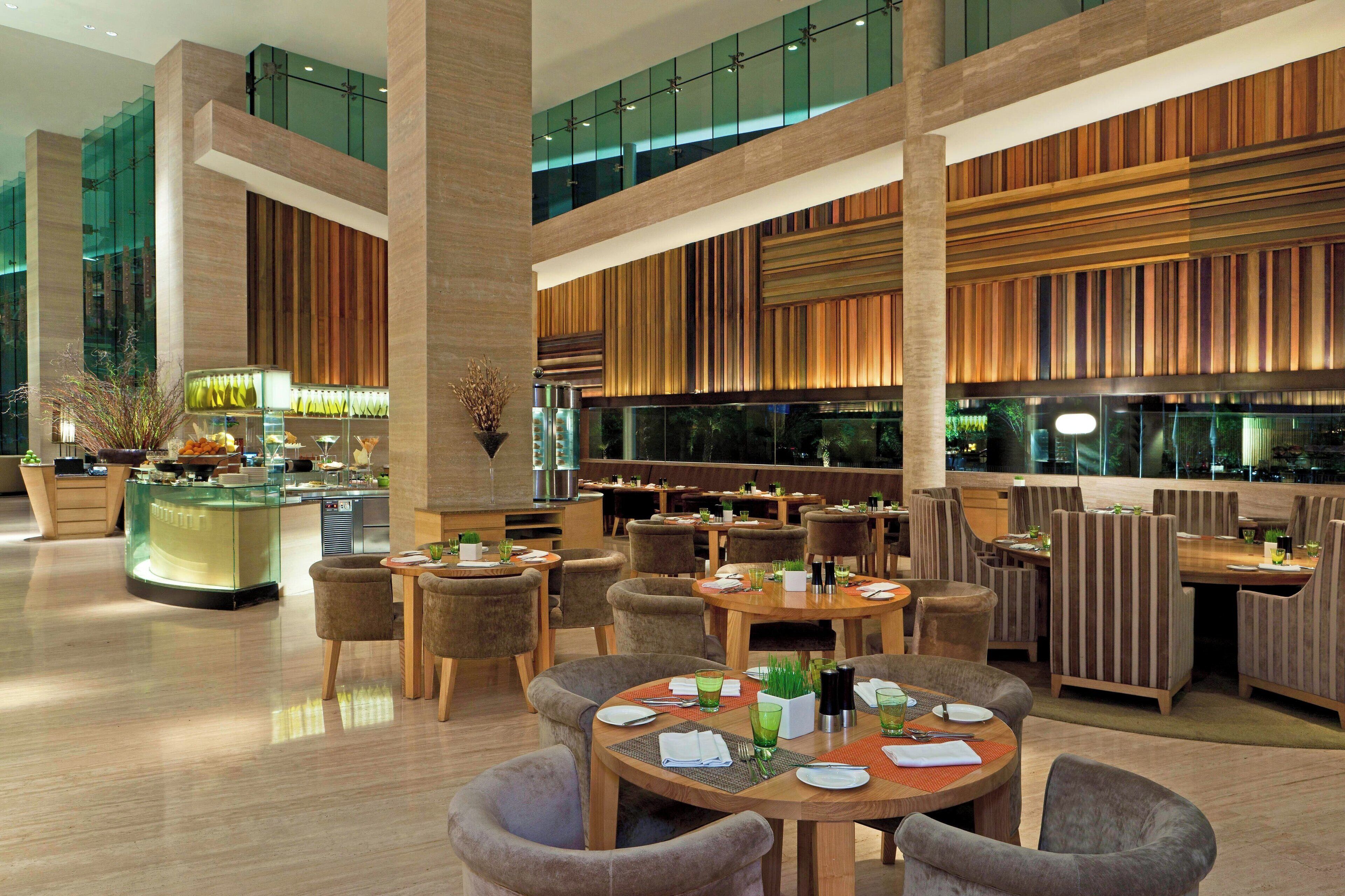 Um amplo hall de hotel com várias áreas de refeições, um bar central e vista para a cidade através de grandes janelas.