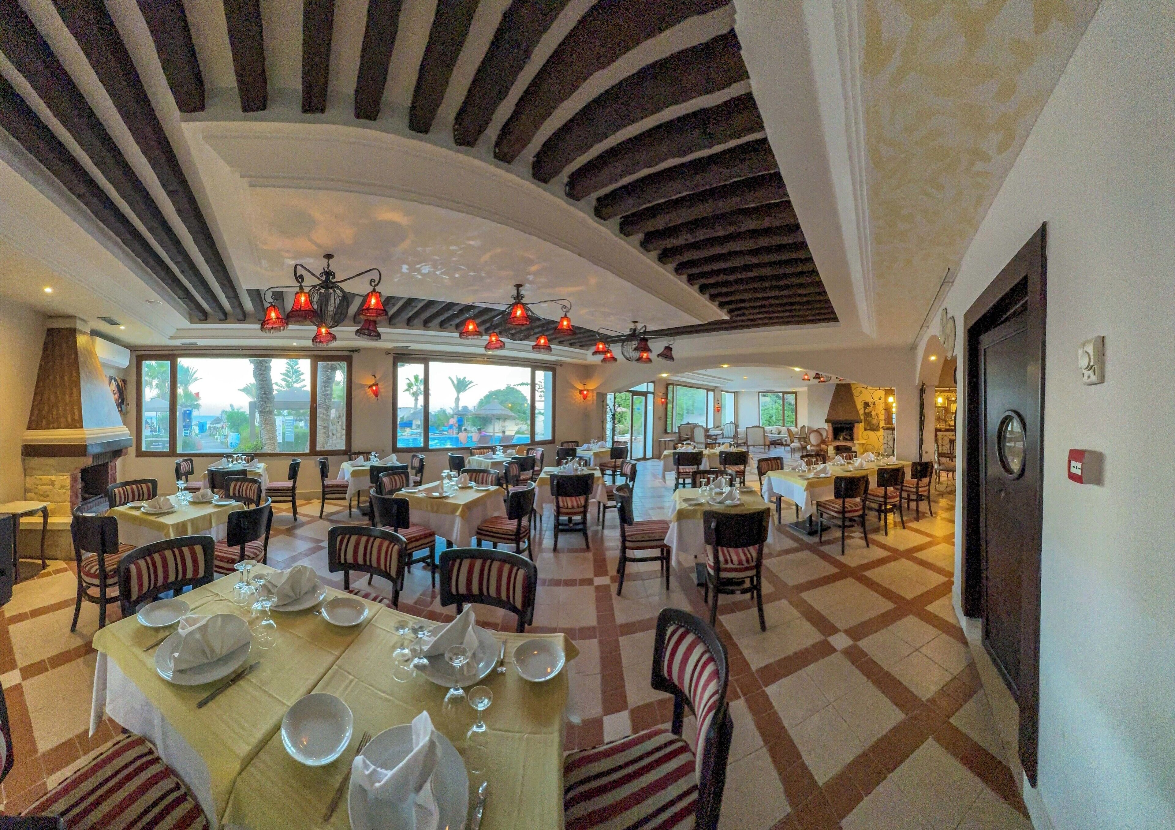 Uma sala de jantar ampla, com várias mesas e cadeiras, janelas grandes com vista e teto alto com vigas aparentes.