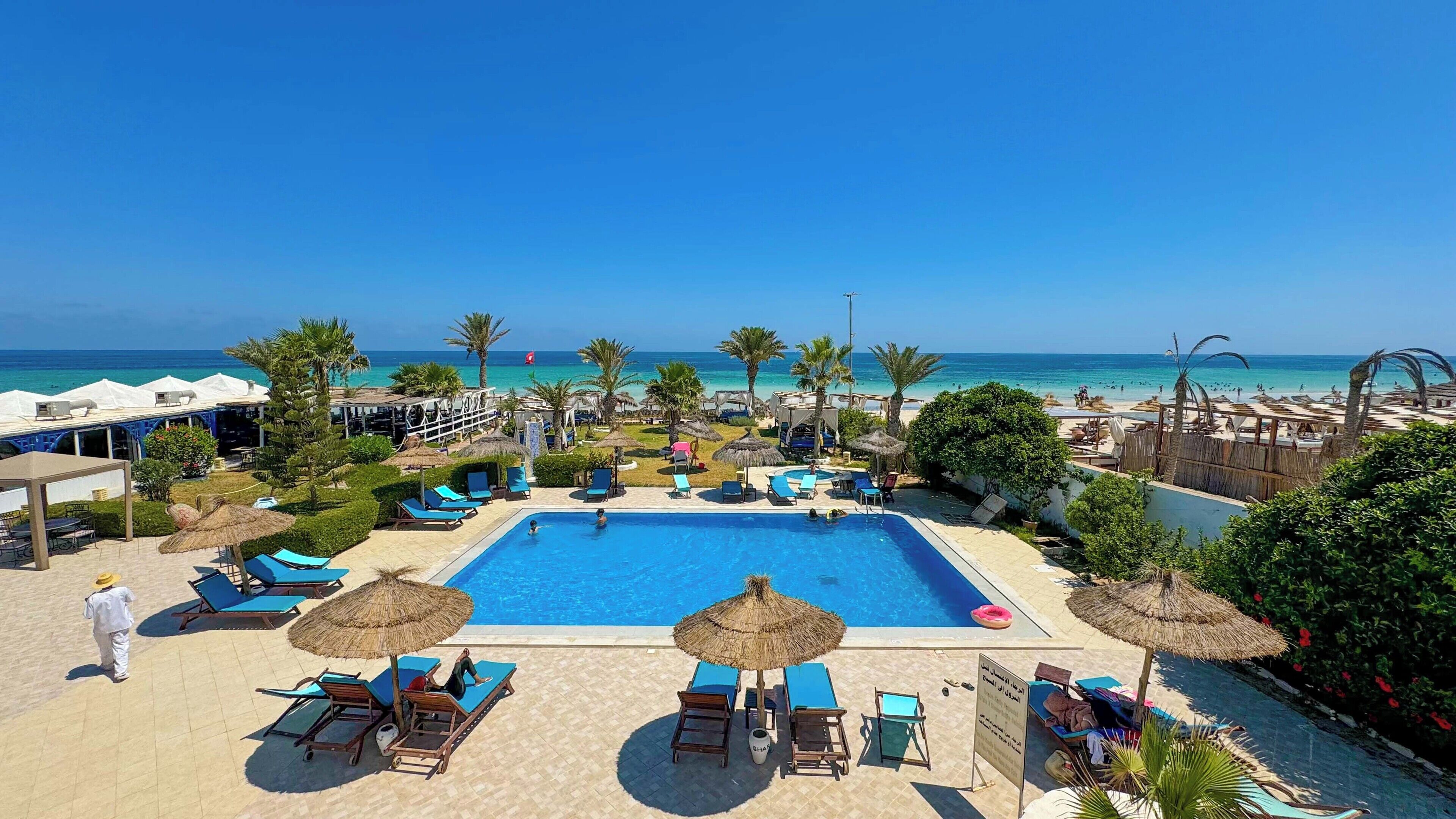 Área da piscina com espreguiçadeiras e guarda-sóis, vista para a praia e céu azul.