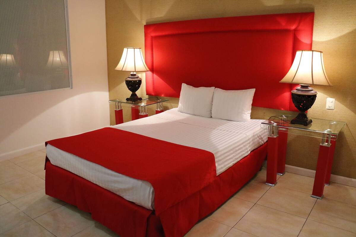 Quarto de hotel com uma cama grande, cabeceira vermelha, duas mesas de cabeceira com luminárias e uma janela com persianas.