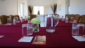 Uma sala de conferências com uma longa mesa posta para uma reunião, incluindo garrafas de água, um jarro e um vaso com flores.