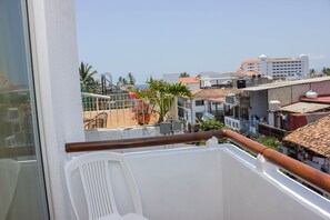 Uma varanda com uma cadeira de plástico branca, guarda-corpo de madeira e vista para uma área residencial com edifícios e palmeiras.