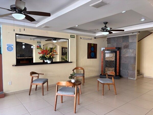 Uma área de recepção com balcão, três cadeiras de madeira com almofadas cinzas, um espelho grande, ventiladores de teto e um painel decorativo na parede.