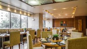 Uma sala de jantar moderna com mesas e cadeiras de madeira, janelas amplas e vista para o verde lá fora.