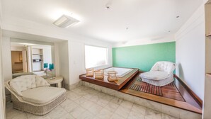Quarto amplo com uma cama grande, uma cadeira e uma mesinha. A parede é verde e há uma janela que permite a entrada de luz natural.