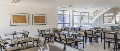 Uma área de refeições de hotel moderna, com várias mesas e cadeiras, janelas amplas e uma escadaria.