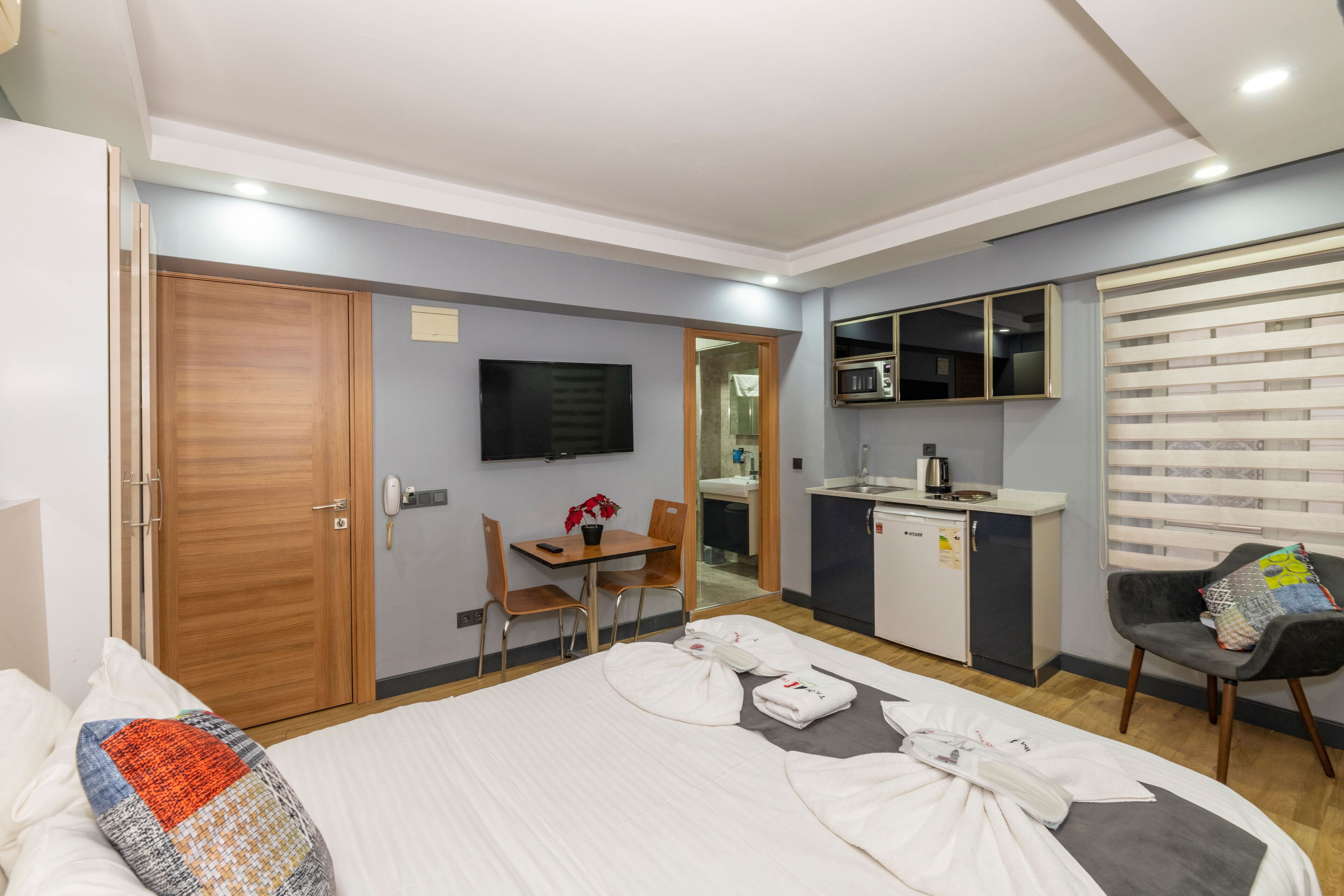 Quarto de hotel com cama, escrivaninha, cadeira, televisão, micro-ondas e pequena cozinha equipada.