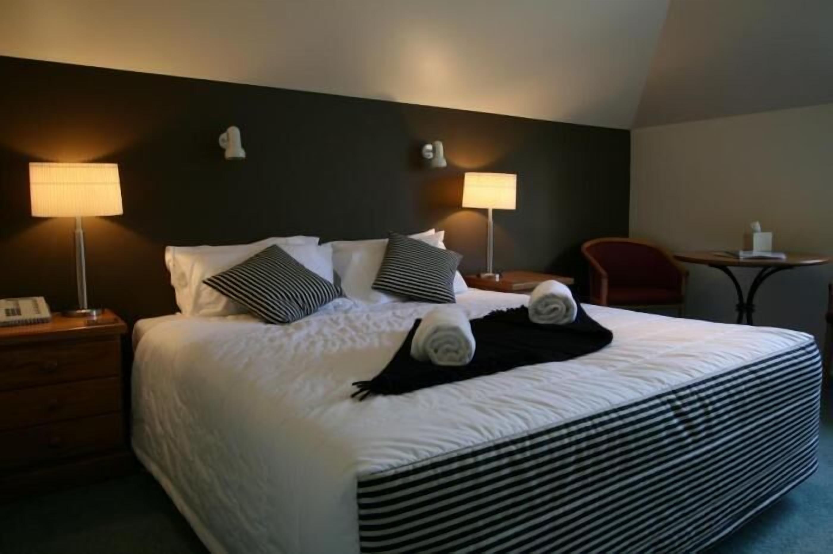 Cama bem arrumada com almofadas listradas, um cobertor preto e toalhas dobradas, ladeada por abajures e uma pequena mesa com um abajur.