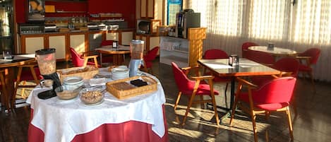 Uma sala de jantar com mesas e cadeiras, um buffet e uma cozinha ao fundo.