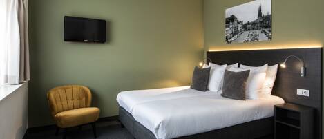 Quarto de hotel com cama, poltrona, televisão e luminária fixa na parede.